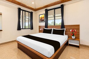 Quarto de hotel com uma cama grande, cabeceira de madeira, duas janelas com cortinas, mesa de cabeceira com um vaso de flores e um quadro na parede.