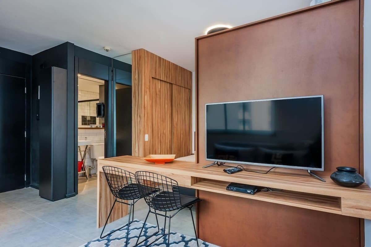 Uma sala de estar moderna com televisão de tela plana, móveis de madeira e uma área de cozinha ao fundo.