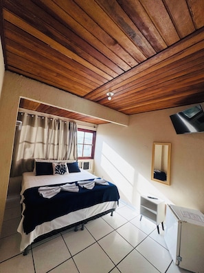 Quarto de hotel com teto de madeira, cama com colcha azul escura, mesa de cabeceira branca, pequeno armário branco, janela com cortinas e televisão fixada na parede.