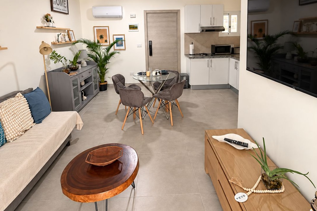 Uma sala de estar moderna com sofá, mesa de centro e área de jantar. Cozinha visível ao fundo, com armários e micro-ondas.