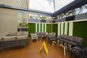Uma área externa com assentos de vime e uma mesa de vidro, rodeada por painéis de grama artificial verde.