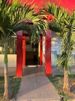Uma entrada de edifício com um arco vermelho, um caminho que leva à entrada e duas palmeiras de cada lado.