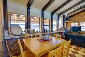 Uma sala de jantar com mesa e cadeiras de madeira, lareira de pedra e grandes janelas com vista para o exterior.