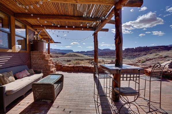 Um pátio externo com pérgola de madeira, luzes de cordão, lareira de tijolos, um sofá e uma mesa com cadeiras.