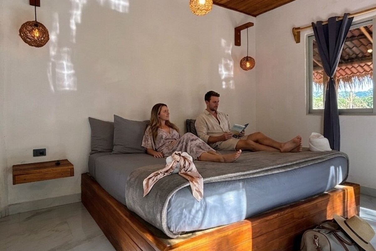 Quarto com teto de madeira, cama com roupa de cama cinzenta e duas pessoas sentadas nela.
