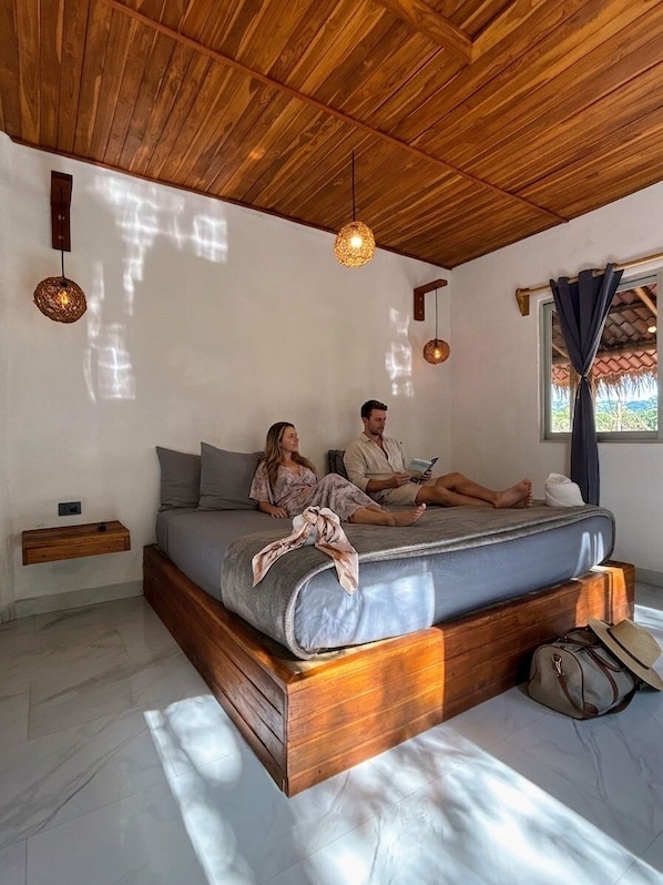 Quarto com teto de madeira, cama com roupa de cama cinzenta e duas pessoas sentadas nela.