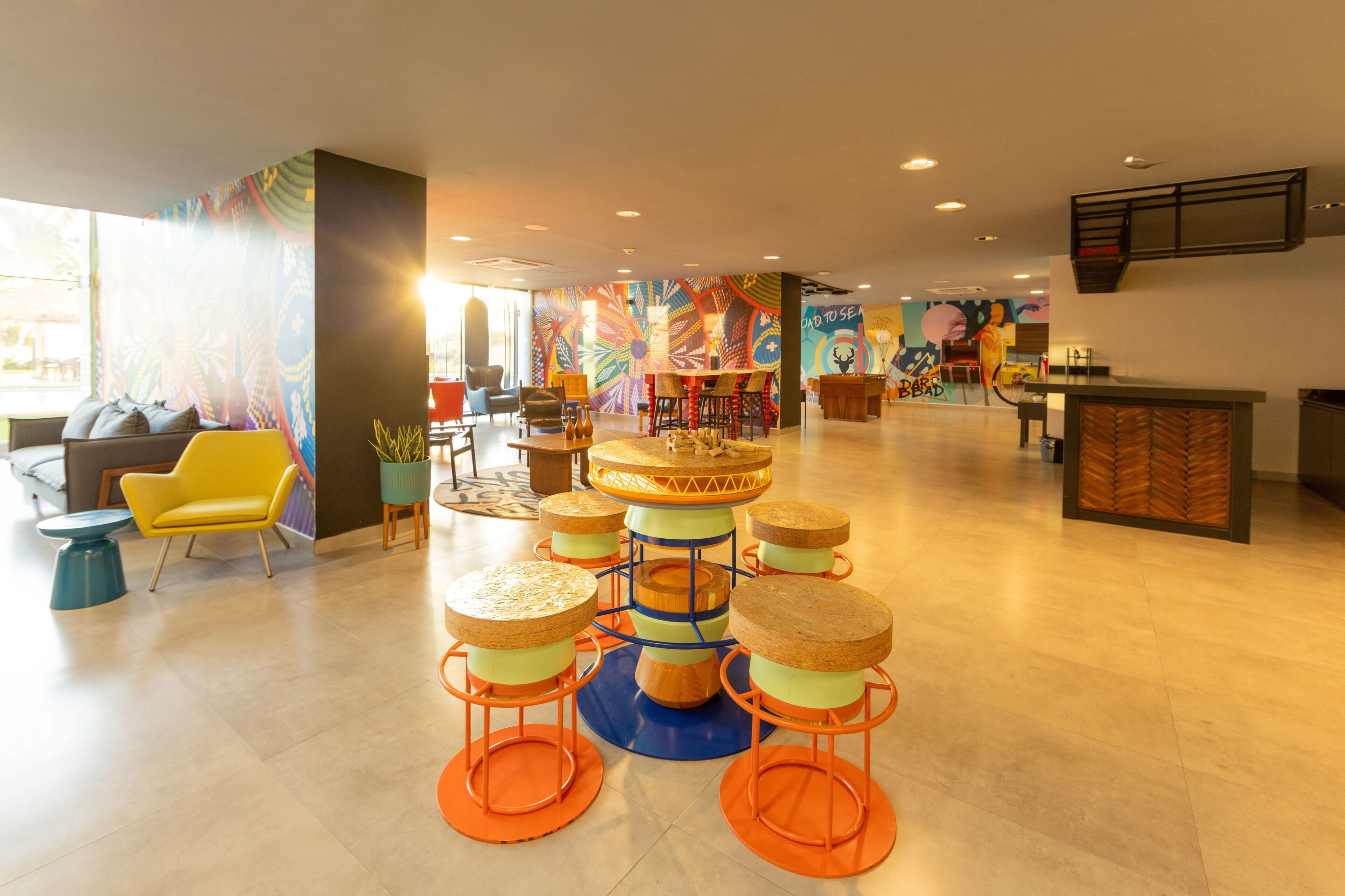 Um lobby moderno com uma mesa redonda central rodeada por banquetas, uma mesa de recepção e obras de arte coloridas nas paredes.