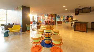Um lobby moderno com uma mesa redonda central rodeada por banquetas, uma mesa de recepção e obras de arte coloridas nas paredes.