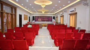 Uma sala de conferências com fileiras de cadeiras vermelhas, um palco com um banner e um candelabro.