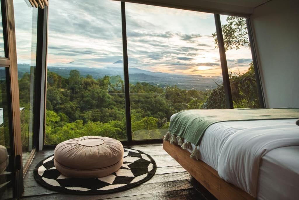 Quarto com uma grande janela, uma cama e um banquinho redondo.