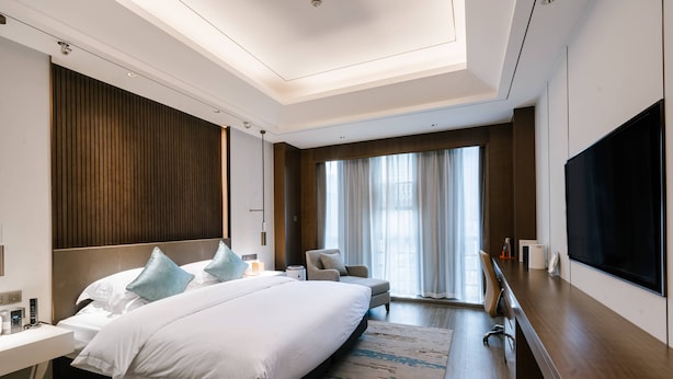 Quarto de hotel moderno com uma cama grande, uma escrivaninha, televisão e uma janela ampla com cortinas.