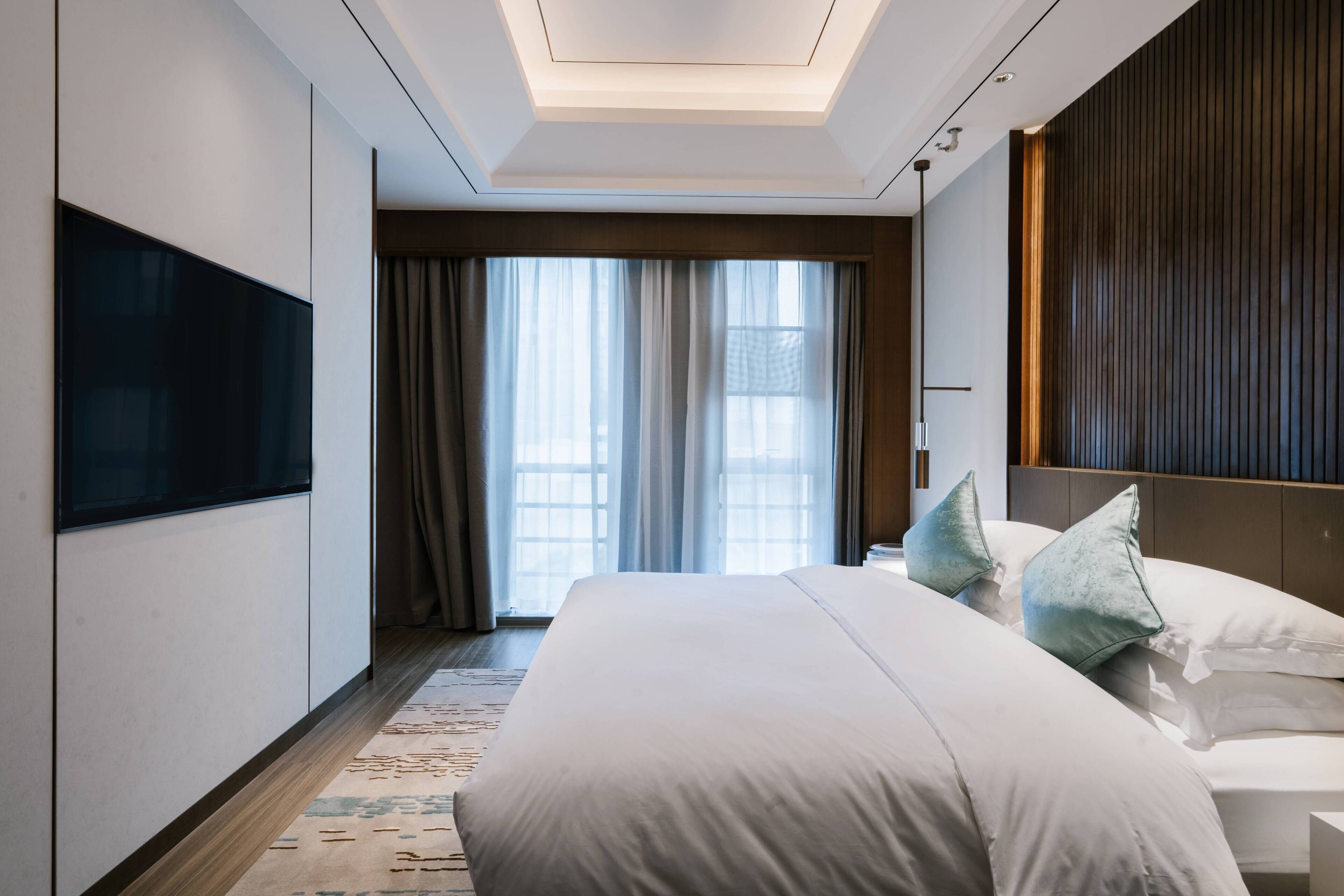 Quarto de hotel moderno com uma cama grande, televisão de tela plana e uma janela ampla com cortinas.