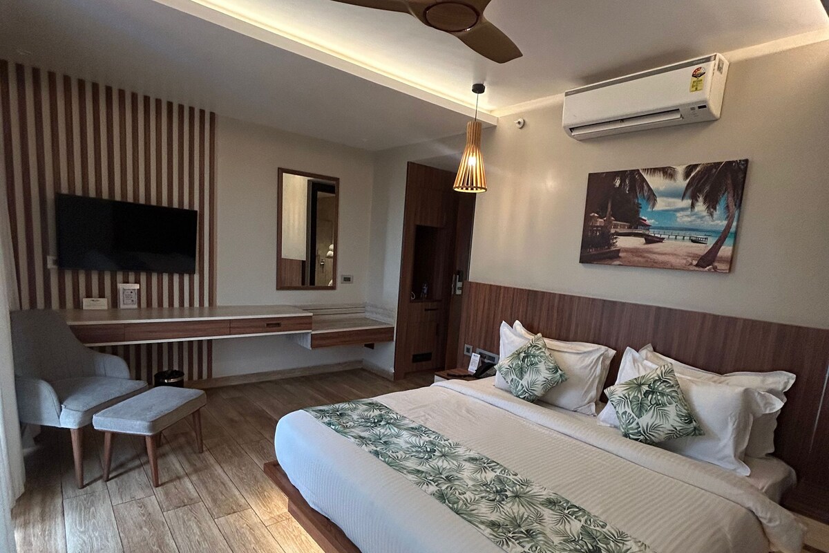 Quarto de hotel com cama grande, escrivaninha, cadeira, ventoinha de teto e ar-condicionado, além de uma pintura na parede.