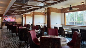 Restaurante com teto alto, vigas de madeira e uma grande tela de televisão. Mesas dispostas com cadeiras vermelhas e toalhas brancas.