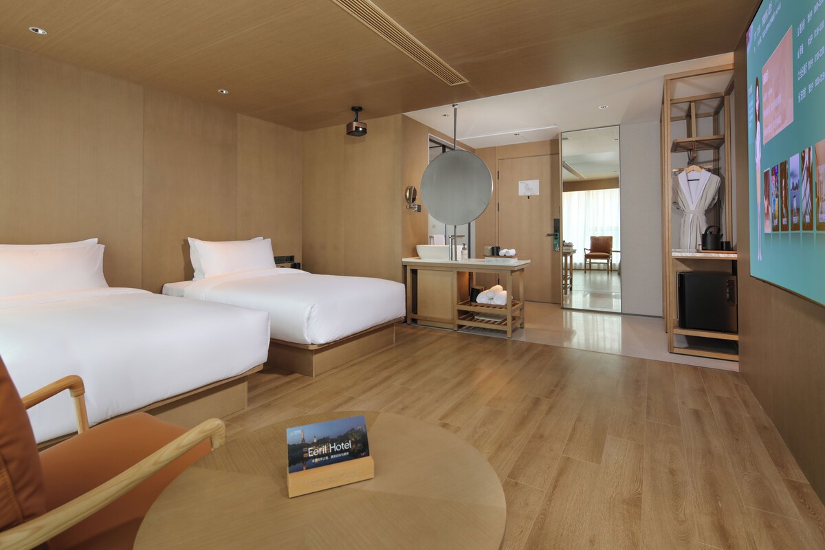 Quarto de hotel com duas camas, uma mesa de escritório com espelho, uma cadeira e uma pequena mesa com um livro.