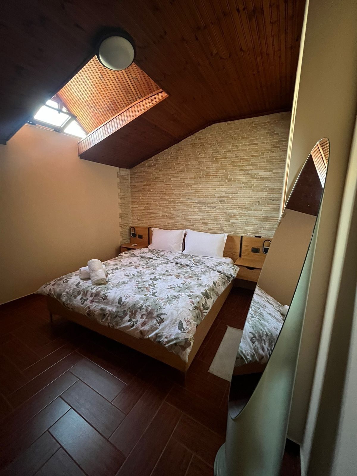 Quarto com teto de madeira, parede de tijolos, cama com roupa de cama florida e um claraboia.