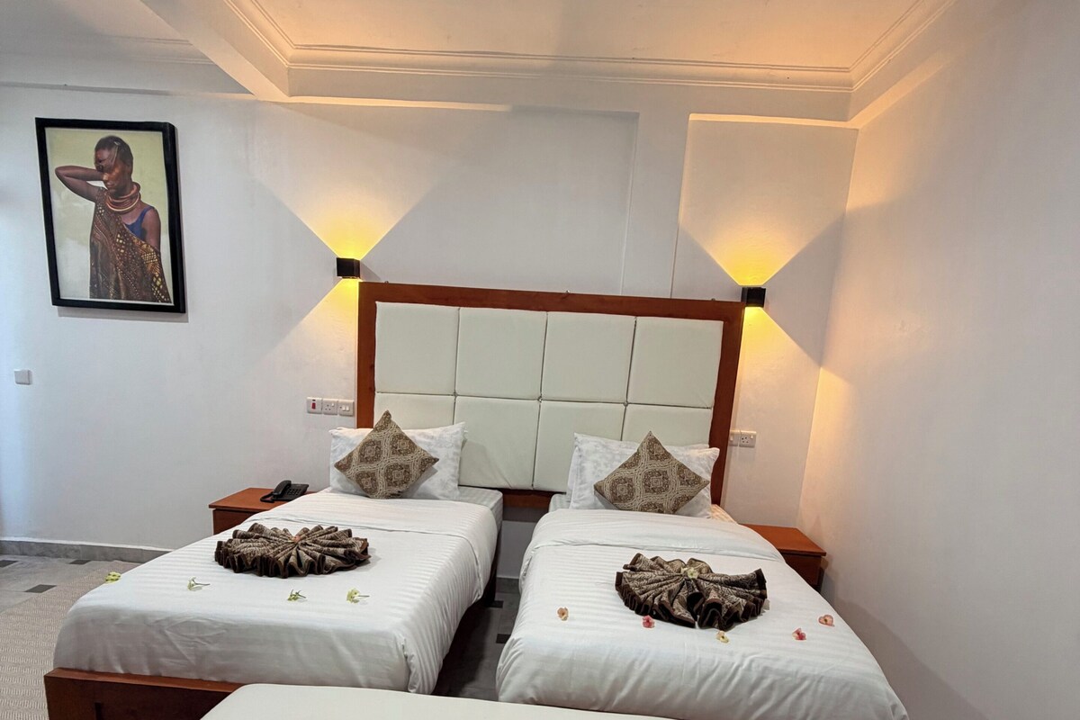 Quarto de hotel com duas camas de solteiro, uma poltrona-sofá maior, cabeceira com painel claro e uma moldura na parede.