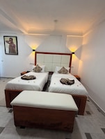 Quarto de hotel com duas camas de solteiro, uma poltrona-sofá maior, cabeceira com painel claro e uma moldura na parede.