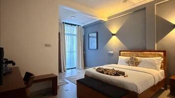 Quarto de hotel com uma cama, mesa de cabeceira e secretária com cadeira. Há uma janela com cortinas e uma porta que dá para outro quarto.