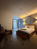 Quarto de hotel com uma cama, mesa de cabeceira e secretária com cadeira. Há uma janela com cortinas e uma porta que dá para outro quarto.