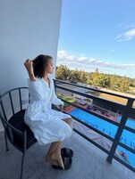 Uma pessoa sentada em uma cadeira de varanda, com vista para uma piscina sob um céu claro e árvores ao fundo.