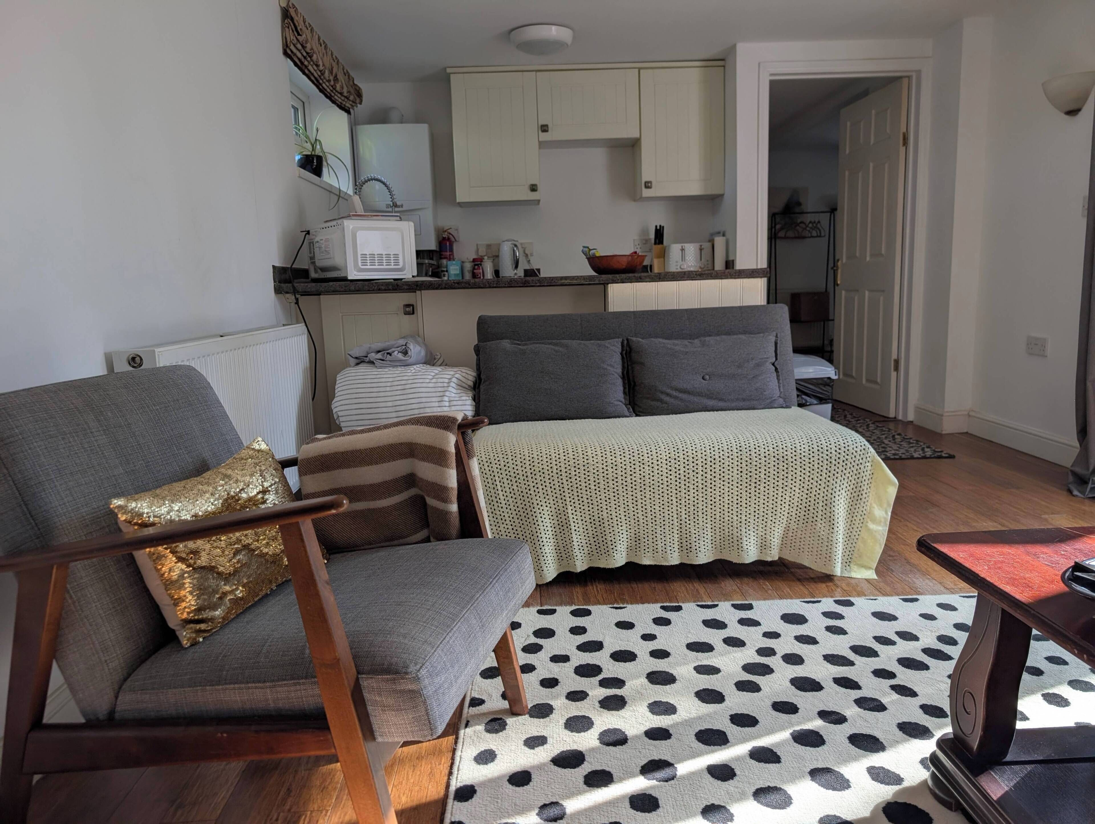 Um espaço compacto com cozinha compacta, uma cama com roupa de cama cinza e branca, uma poltrona cinza com almofada dourada e um tapete com bolinhas.