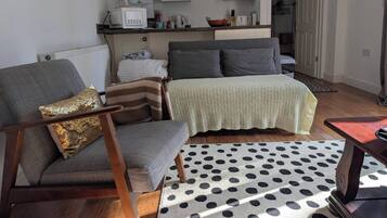 Um espaço compacto com cozinha compacta, uma cama com roupa de cama cinza e branca, uma poltrona cinza com almofada dourada e um tapete com bolinhas.