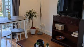 Uma sala de estar com televisão sobre um móvel, uma mesa de jantar com cadeiras, um vaso de planta e uma estante de livros.