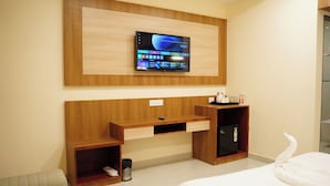 Quarto de hotel com televisão de tela plana na parede, uma escrivaninha de madeira com gaveta, uma pequena lixeira e uma cama com travesseiro branco.