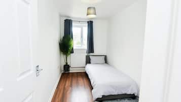 Quarto pequeno e limpo, com uma cama, radiador, janela com cortinas e uma planta.