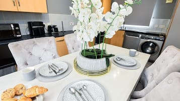 Uma cozinha moderna com uma mesa de jantar posta para dois, com um vaso de orquídeas brancas, uma cafeteira e uma máquina de lavar roupa ao fundo.