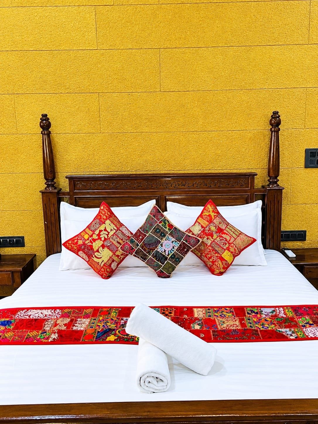 Cama com cabeceira de madeira, duas almofadas vermelhas, uma almofada estampada e uma toalha branca enrolada em um cobreleite vermelho e branco.