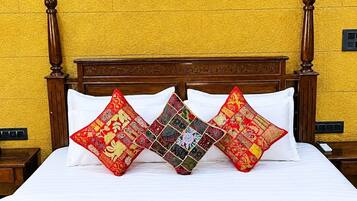 Cama com cabeceira de madeira, duas almofadas vermelhas, uma almofada estampada e uma toalha branca enrolada em um cobreleite vermelho e branco.