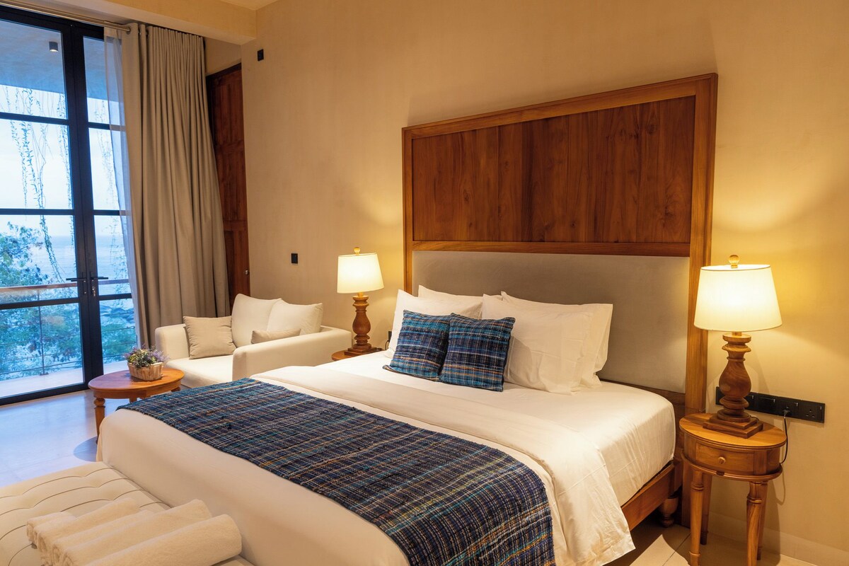 Quarto de hotel com uma cama grande, duas mesas de cabeceira com abajures, uma janela com cortinas e vista para o exterior.