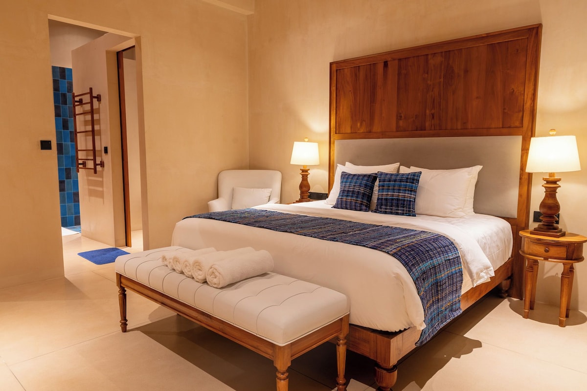 Quarto de hotel com cama grande, duas mesinhas de cabeceira, um banco e cabeceira de madeira.