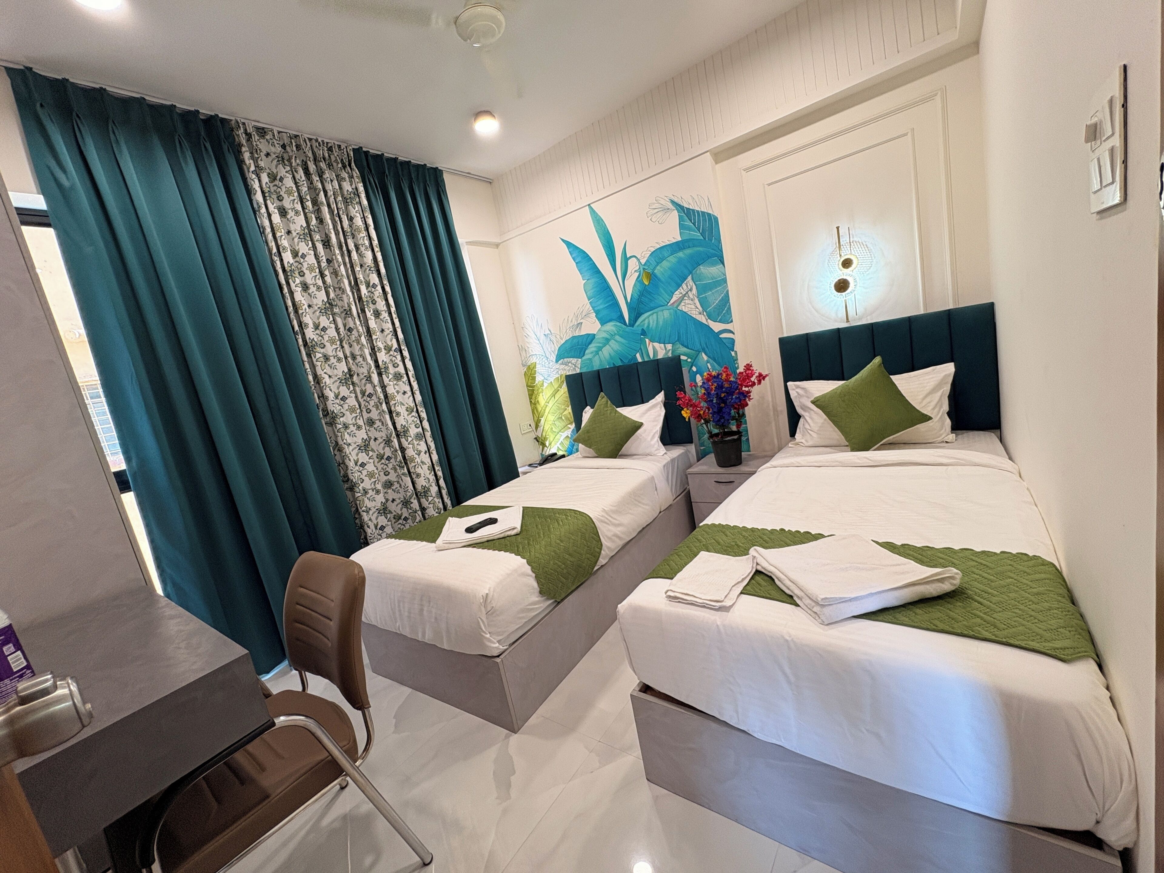 Quarto de hotel com duas camas, uma escrivaninha, uma cadeira e uma janela grande com cortinas.