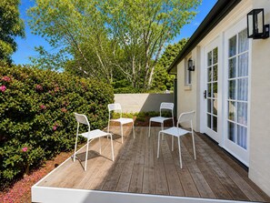 Um deck de madeira com quatro cadeiras brancas e uma mesa branca, cercado por vegetação exuberante e um prédio branco com janelas grandes.