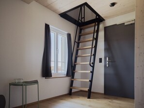 Quarto com escada de madeira, janela com cortinas, porta e pequena mesa com copos.
