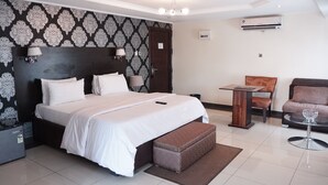 A hotel room with a large bed, bedside lamps, a small table, a chair, and an air conditioning unit on the wall.