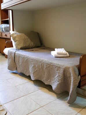 Quarto pequeno com uma cama de solteiro, colcha acolchoada, toalha branca dobrada e mesa de cabeceira de madeira com abajur.