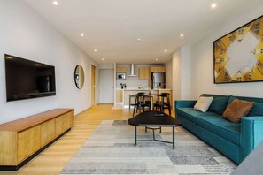 Uma sala de estar moderna com sofá, mesa de centro, televisão e cozinha ao fundo.