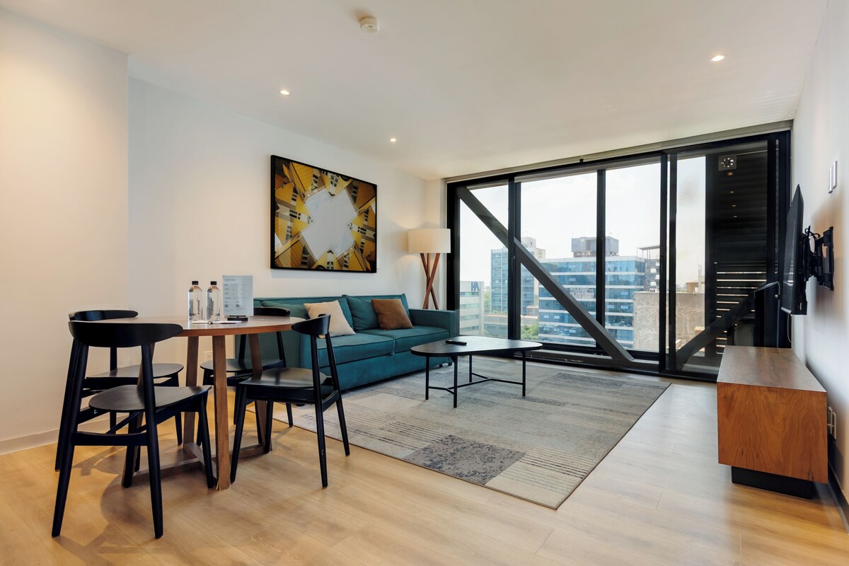 Uma sala de estar moderna com sofá, mesa de centro e cadeiras, uma janela grande com vista para a cidade e um quadro na parede.