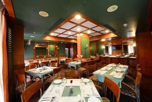 Restaurante com várias mesas e cadeiras, teto alto com iluminação embutida e área de cozinha visível ao fundo.