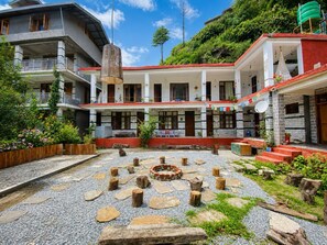 Um pátio com lareira central rodeada por lajes de pedra e tocos de madeira, ladeado por edifícios com varandas e plantas em vasos.