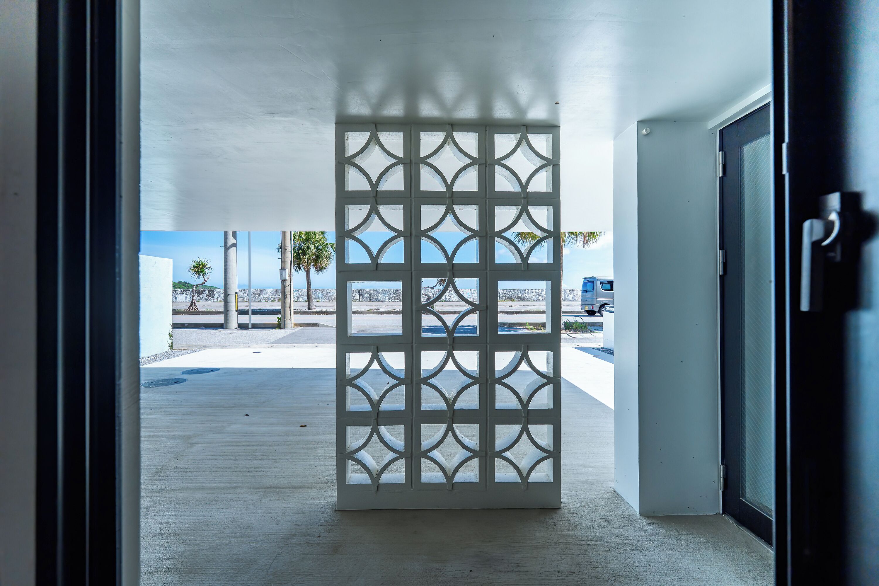 Visão através de uma porta com um painel decorativo, dando para uma área externa com piso de concreto e céu claro.