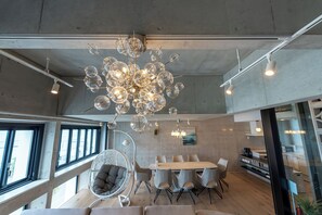 Um interior moderno com um lustre esférico único, uma área de refeições com mesa e cadeiras redondas, e uma cadeira de vime com almofada.