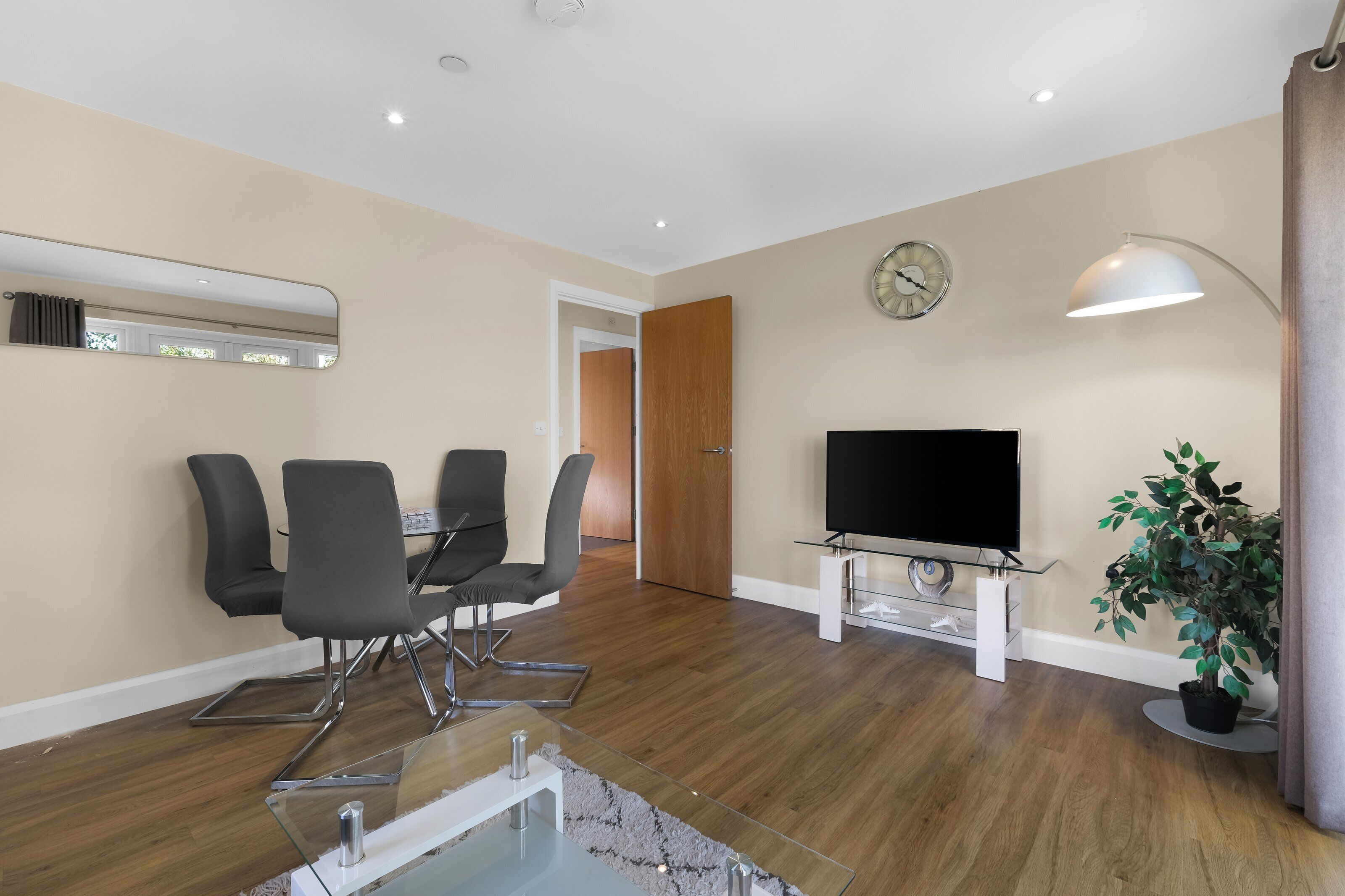 Uma sala moderna com televisão de tela plana, área de jantar com cadeiras, mesa de centro de vidro e um vaso com planta.