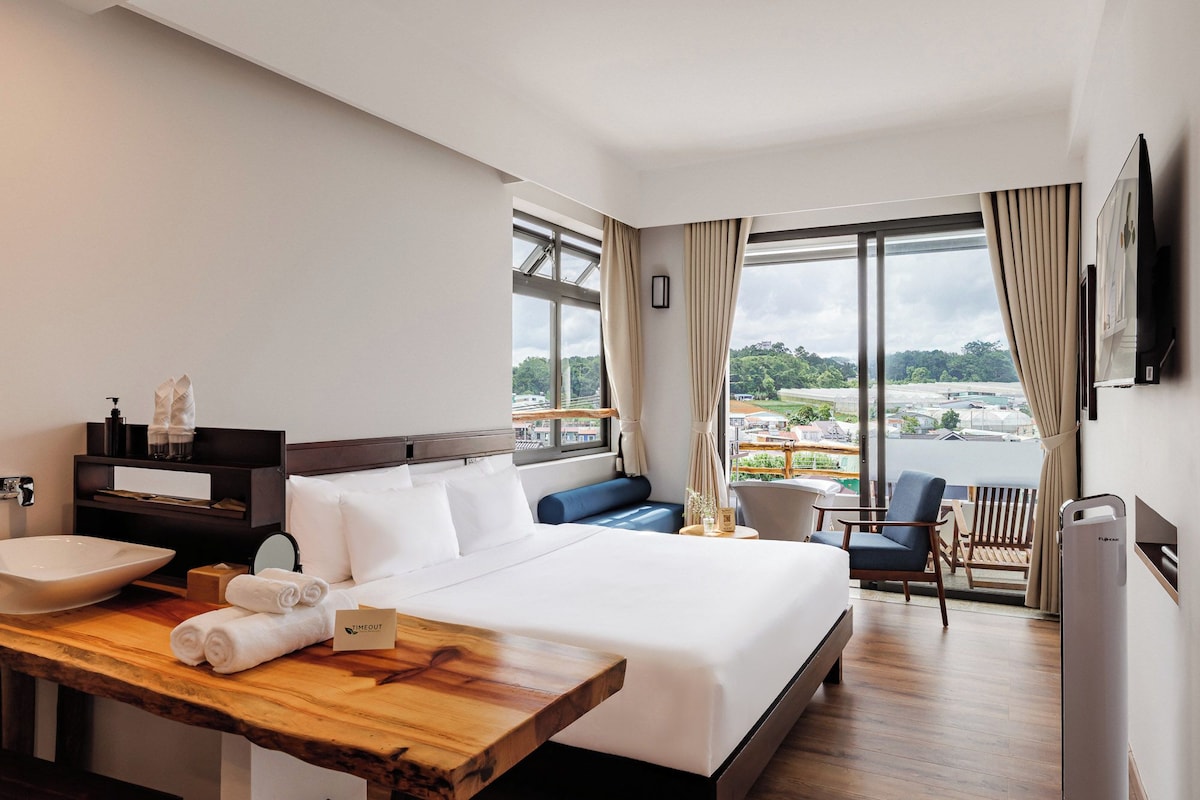 Quarto de hotel com uma cama, uma mesa de madeira com toalhas, pia e vista para a cidade através de grandes janelas.