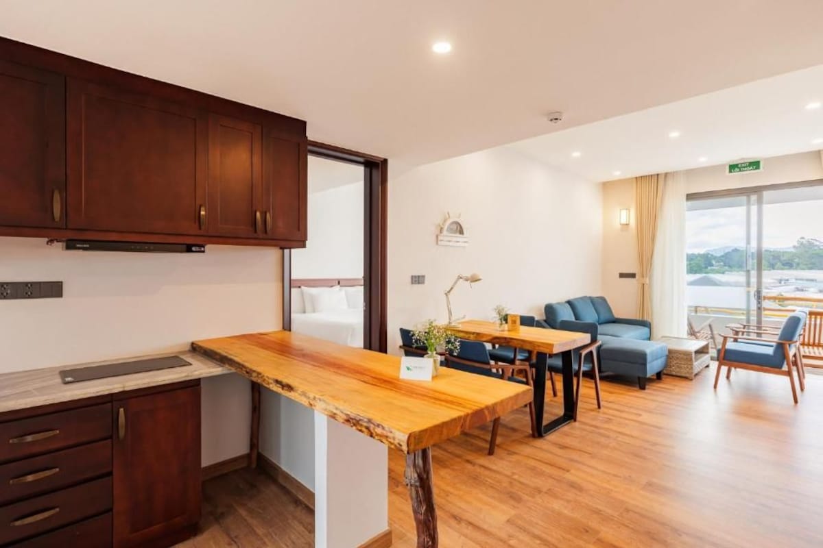 Cozinha moderna com armários e bancada de madeira. Há uma área de jantar com mesa e cadeiras, e um sofá. O espaço possui uma janela grande com vista.