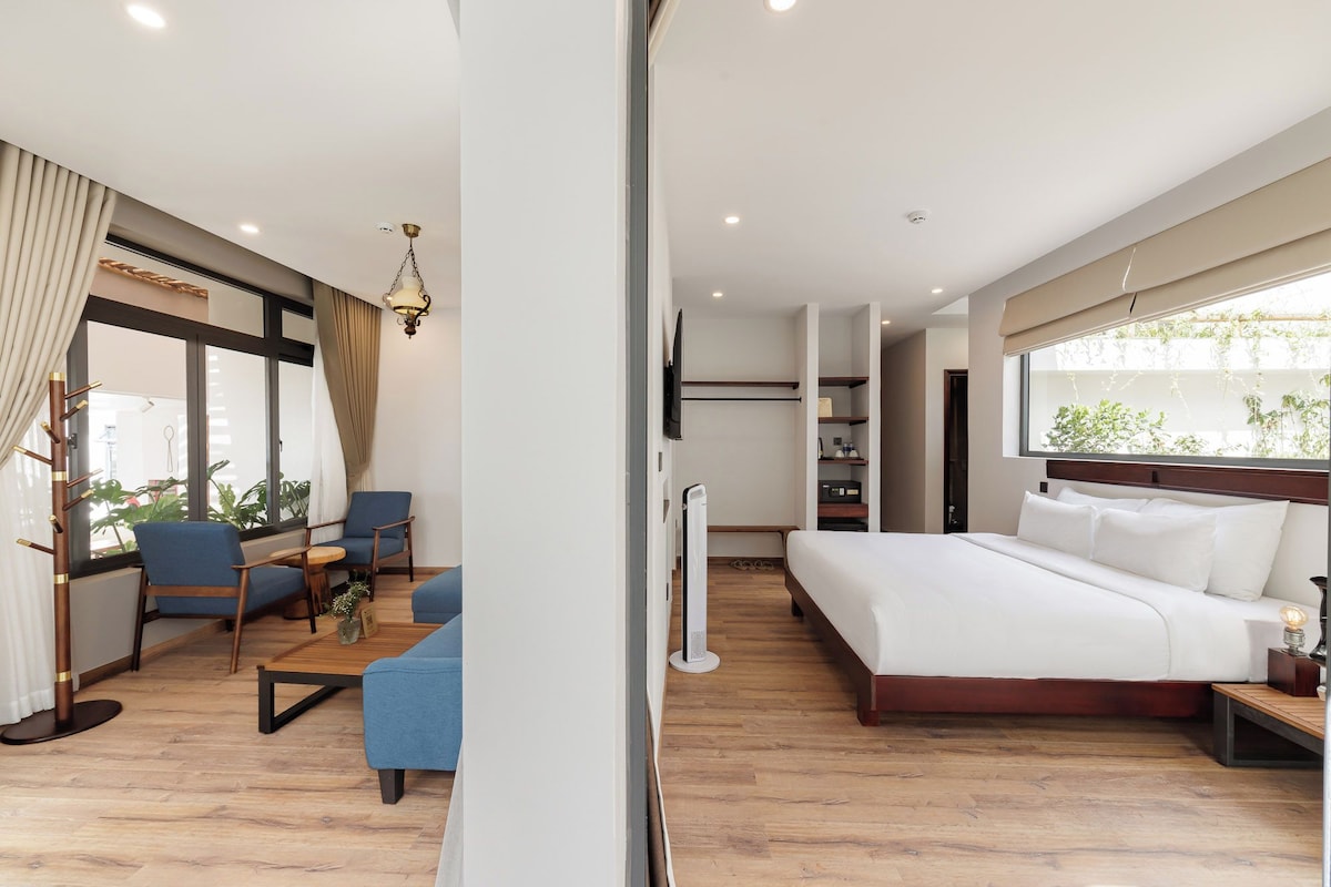 Quarto de hotel moderno com uma cama grande, área de estar com cadeiras, televisão e janela com vista para o verde.